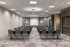 Uma sala de conferências com fileiras de mesas e cadeiras, uma tela grande na frente e um design moderno no teto.