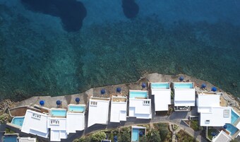Vista aérea de um resort à beira-mar com vários edifícios brancos, cada um com piscina no terraço e guarda-sóis azuis.