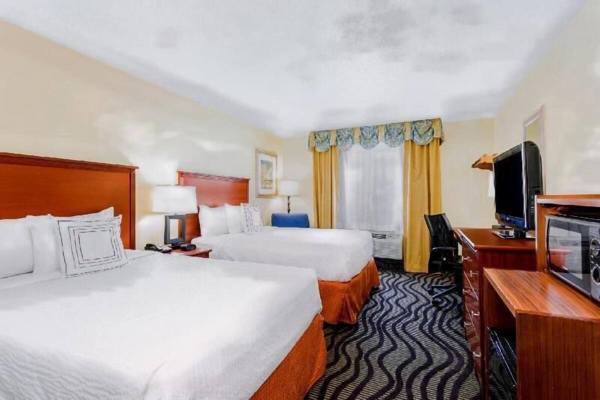 Quarto de hotel com duas camas, uma escrivaninha com televisão, uma lâmpada e uma cadeira.