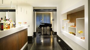 Um hall de hotel moderno com piso preto polido, um piano e uma área de bar com diversos frascos e copos.