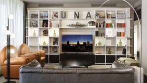 Uma sala de estar moderna com televisão exibindo uma paisagem urbana, sofás de couro, uma estante com livros e objetos decorativos, e um abajur.