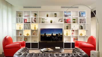 Uma sala de estar moderna com uma TV de tela plana, poltronas de couro vermelho, um banquinho de couro preto e uma estante branca com diversos objetos decorativos.