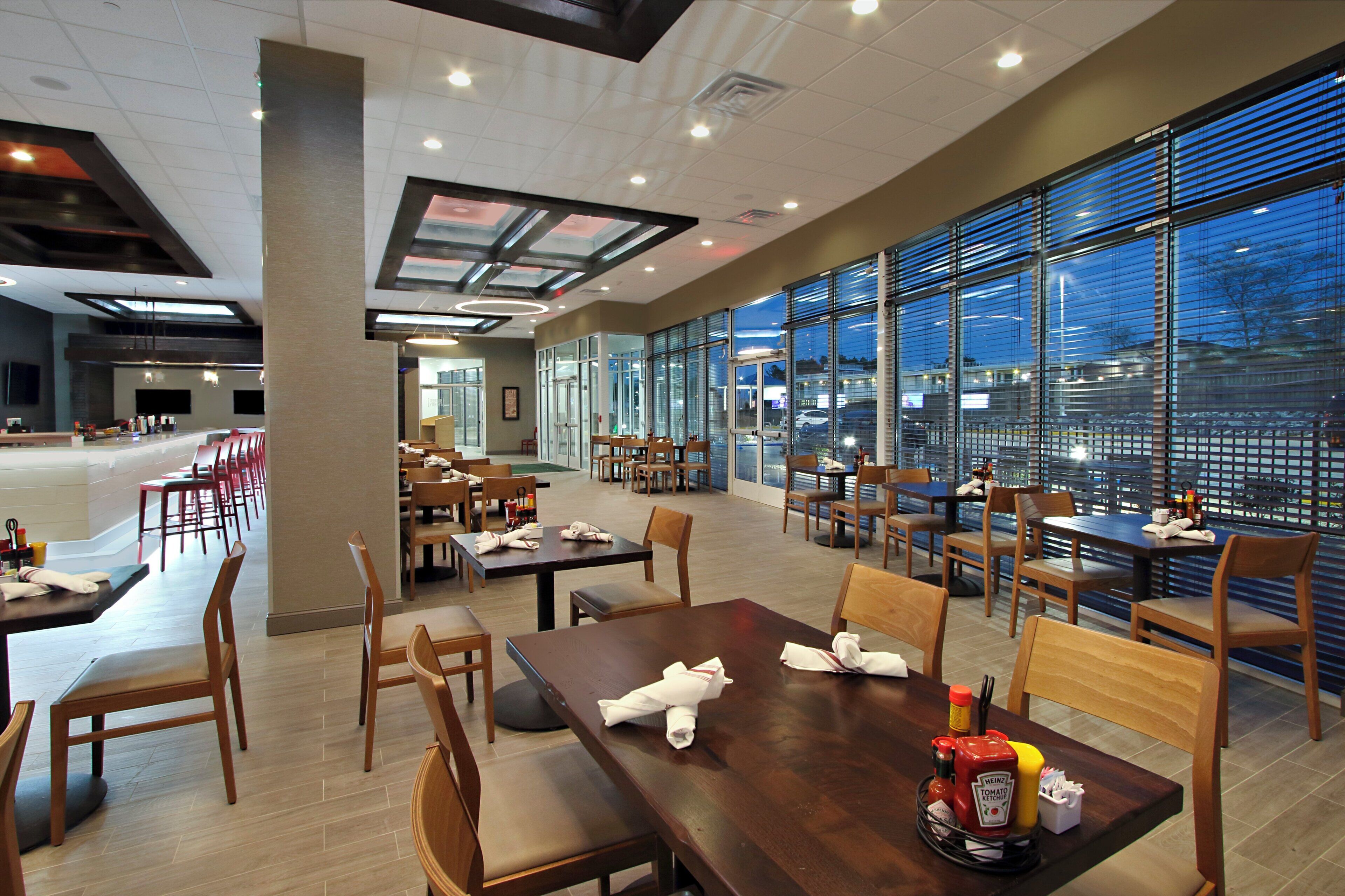 Um restaurante amplo com várias mesas e cadeiras, janelas grandes que oferecem vista para o exterior e um design moderno no teto.