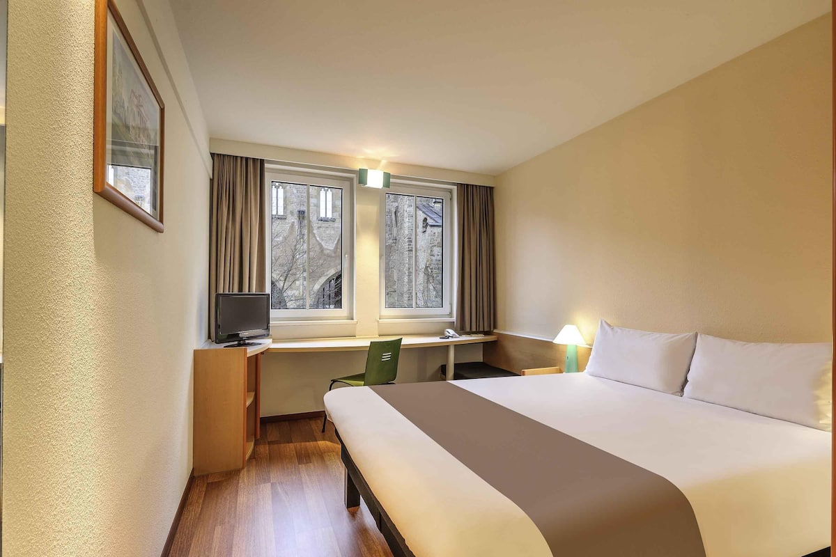 Quarto de hotel com cama, escrivaninha, cadeira e janela com cortinas.