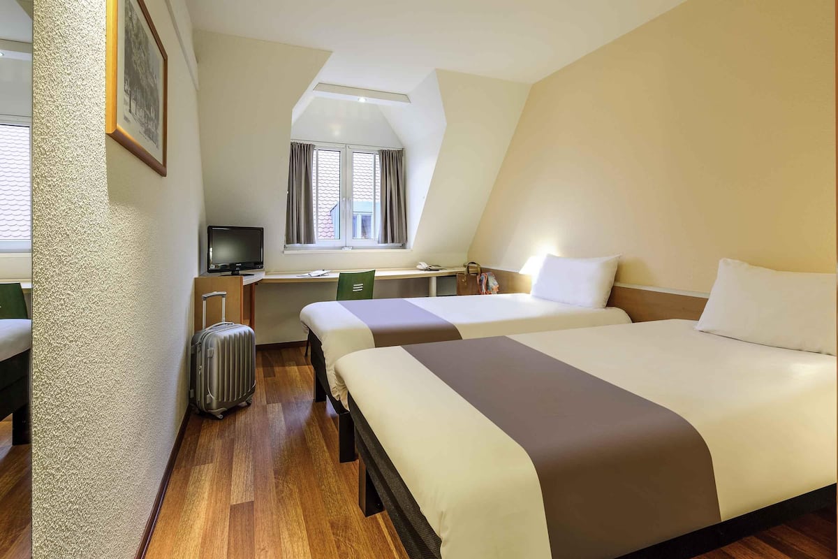 Quarto de hotel com duas camas, uma escrivaninha, uma cadeira, uma televisão e uma janela com cortinas.