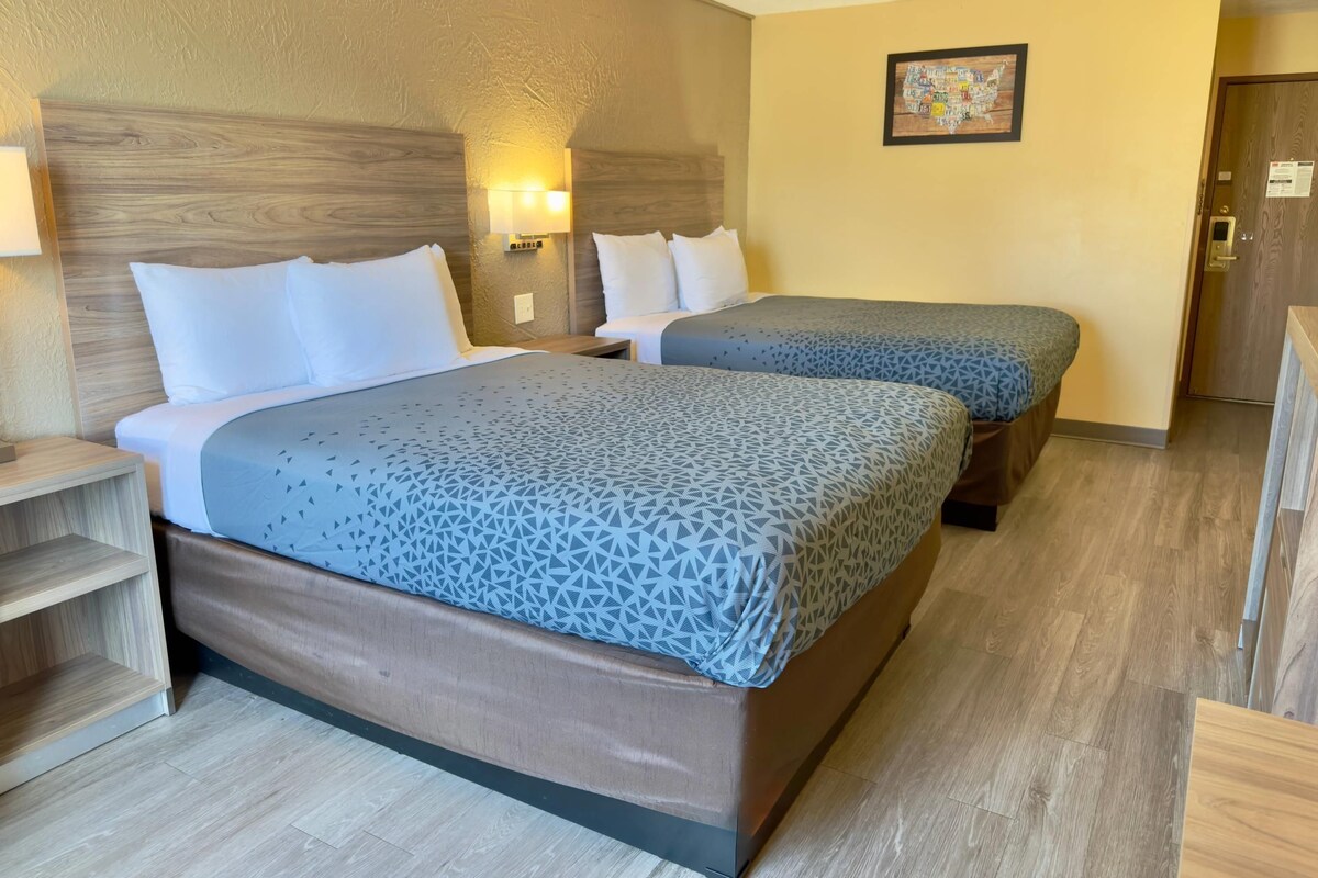 Quarto de hotel com duas camas, cabeceira de madeira, mesa de cabeceira com abajur e luminária na parede.