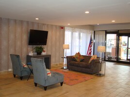Um hall de hotel com um sofá cinzento, duas poltronas, uma televisão de tela plana, um vaso de planta e uma bandeira americana.