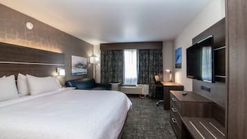 Quarto de hotel com uma cama grande, uma escrivaninha com televisão de tela plana, uma cadeira, abajur e uma janela com cortinas.