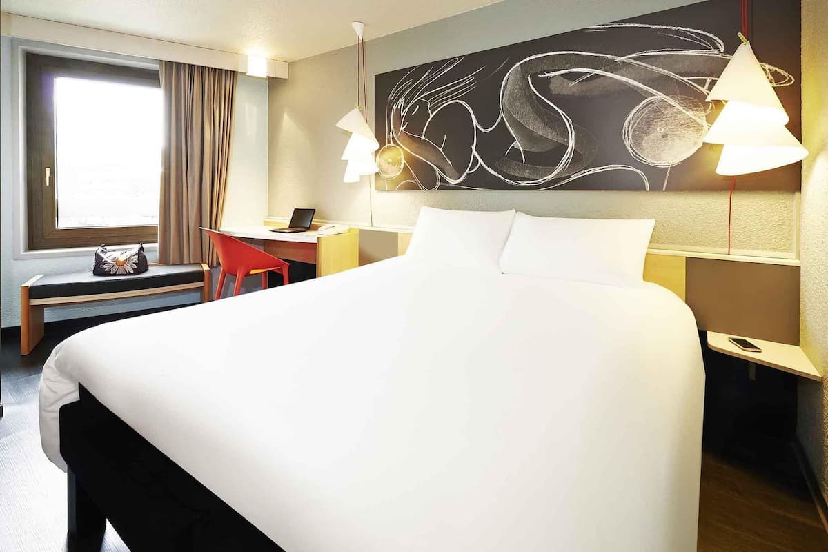 Quarto de hotel com uma cama grande, uma escrivaninha, uma cadeira, janela com cortinas e uma obra de arte na parede.
