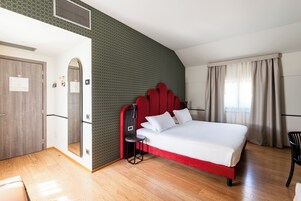 Quarto com cabeceira vermelha, piso de madeira, cama com roupa de cama branca, uma pequena mesa e uma cadeira.