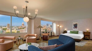 Quarto de hotel moderno com uma grande janela com vista para a cidade, um candelabro, uma cama com travesseiro verde e um sofá azul.