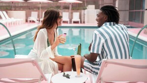 Duas pessoas sentadas à mesa junto à piscina, com uma bebida, rodeadas por espreguiçadeiras e guarda-sóis.