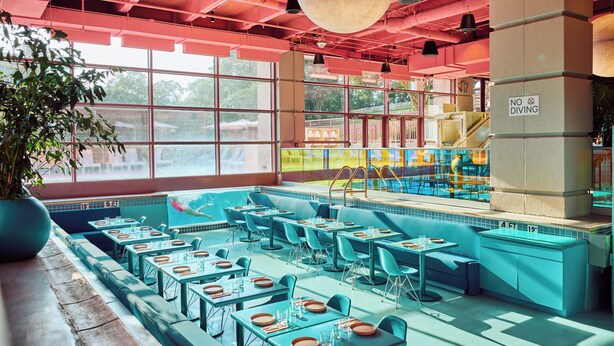 Restaurante à beira da piscina com mesas e cadeiras, uma grande planta em vaso e um aviso de 'No Diving'.