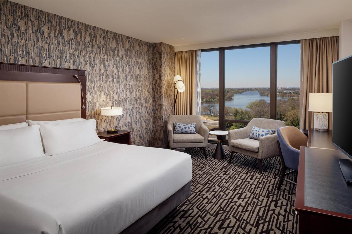 Quarto de hotel com uma cama grande, duas poltronas, uma televisão e vista para um lago.