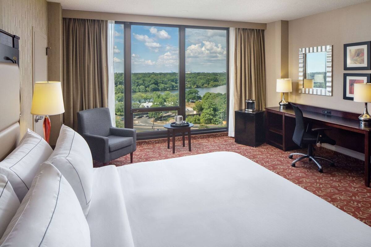 Quarto de hotel com uma cama grande, uma escrivaninha, uma cadeira, uma mesa pequena e vista para um lago e árvores.