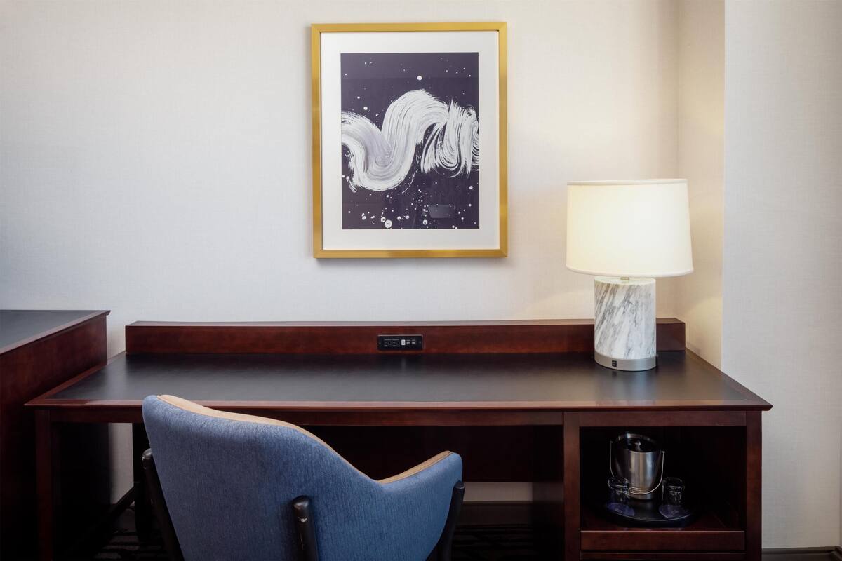 Quarto de hotel com uma escrivaninha, uma cadeira, uma obra de arte emoldurada, um abajur e uma cafeteira.