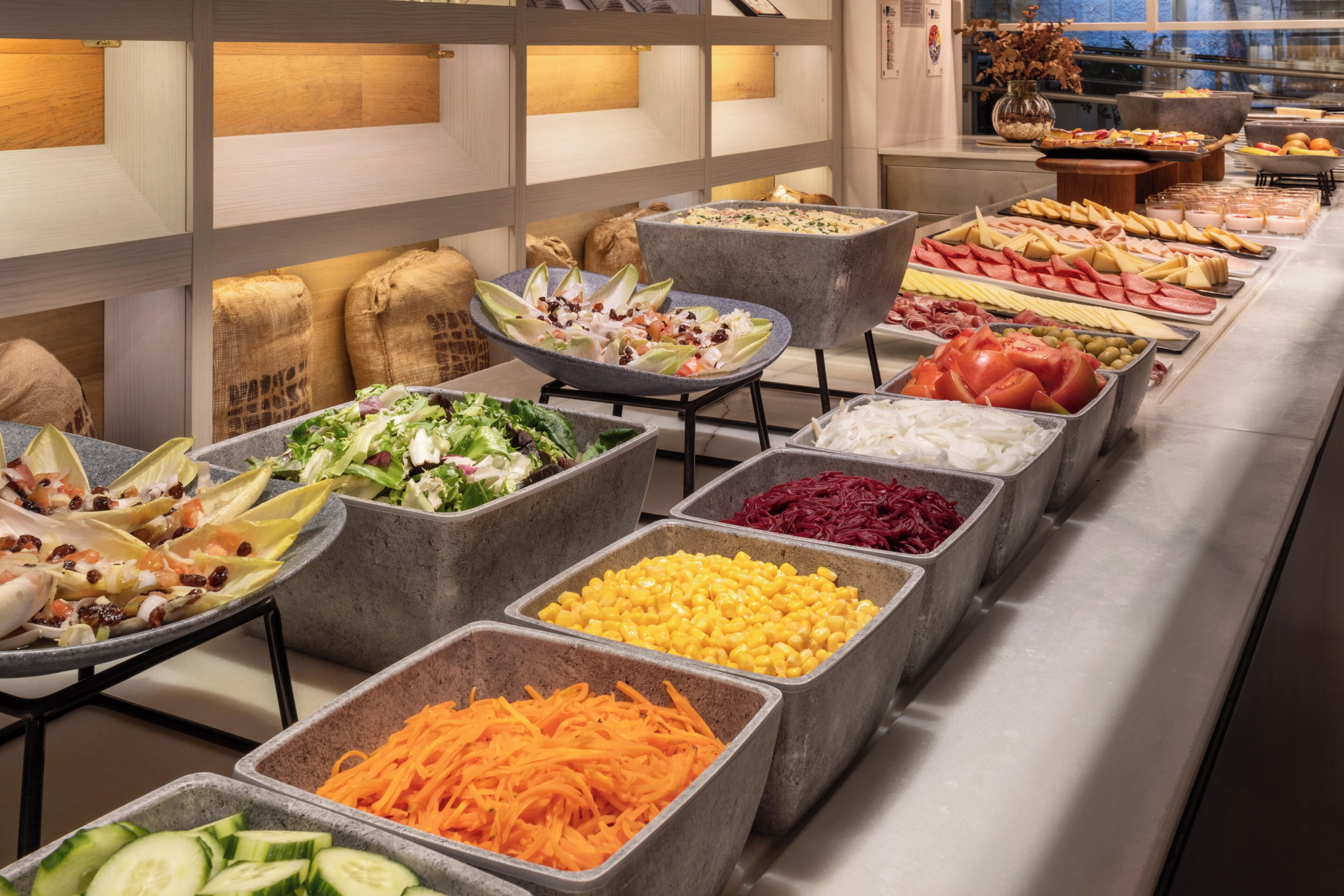Buffet com diversos alimentos em recipientes de metal, incluindo saladas, frutas e legumes.