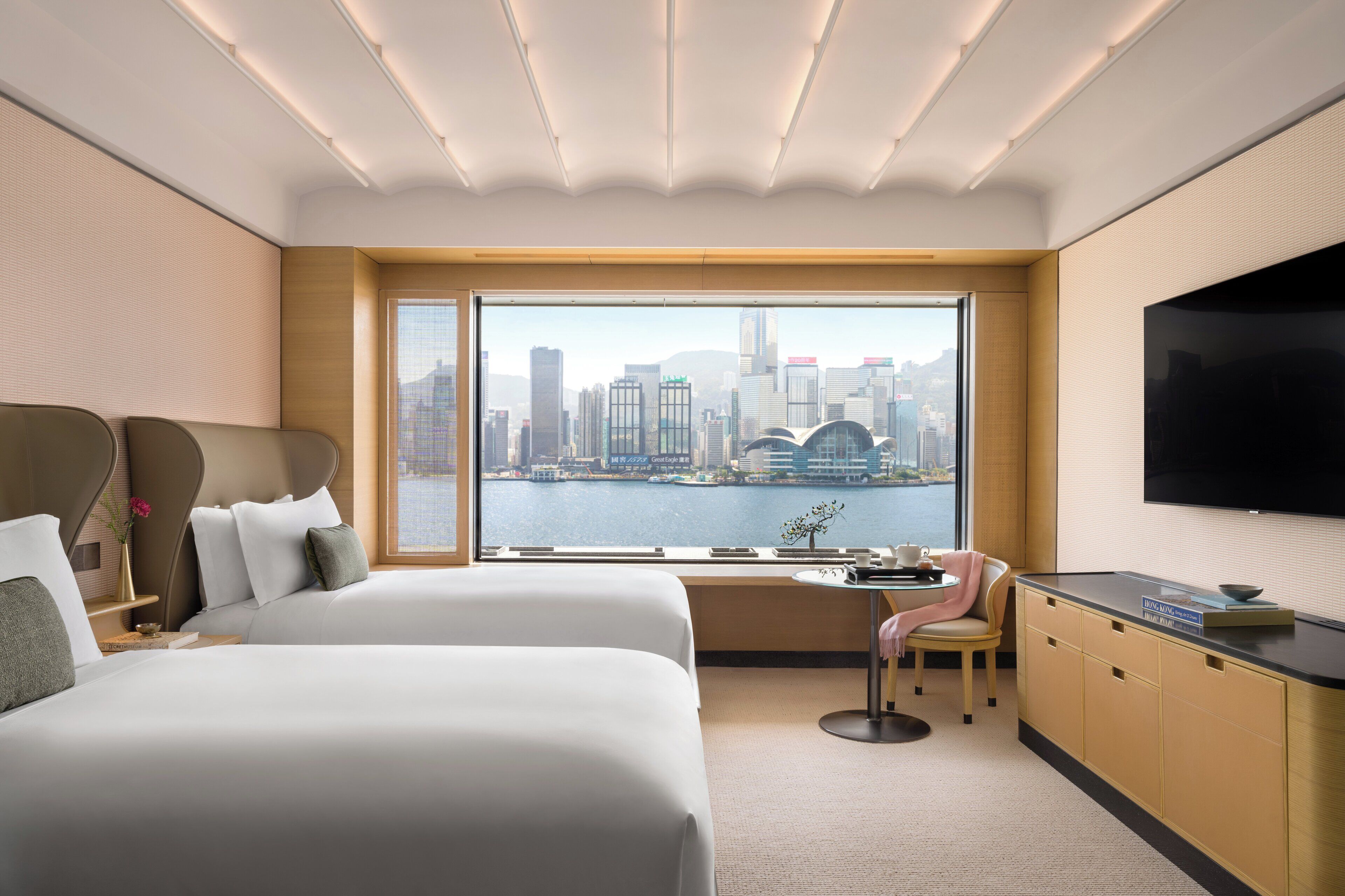 Quarto de hotel com duas camas, uma escrivaninha, uma cadeira, televisão e uma janela grande com vista para a cidade.