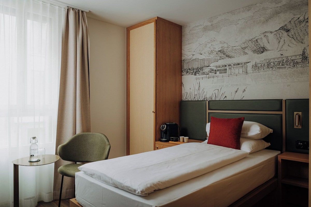 Quarto de hotel com cama, cadeira, mesa pequena, máquina de café e parede com pintura de paisagem.