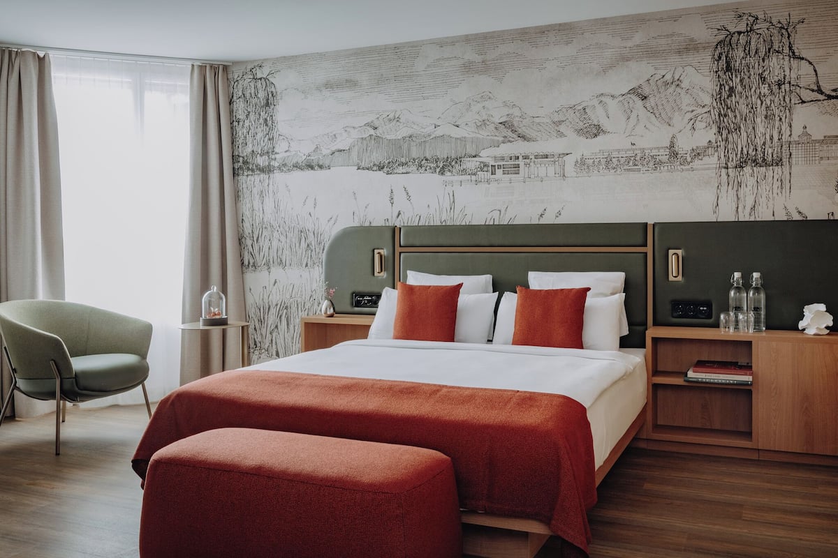 Quarto de hotel com uma cama grande, uma poltrona verde, uma mesa de cabeceira com abajur e um mural na parede.