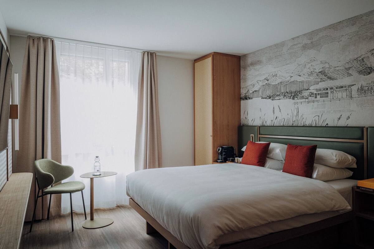 Quarto de hotel com cama, uma cadeira, uma mesinha com uma garrafa, um quadro na parede e uma janela com cortinas.