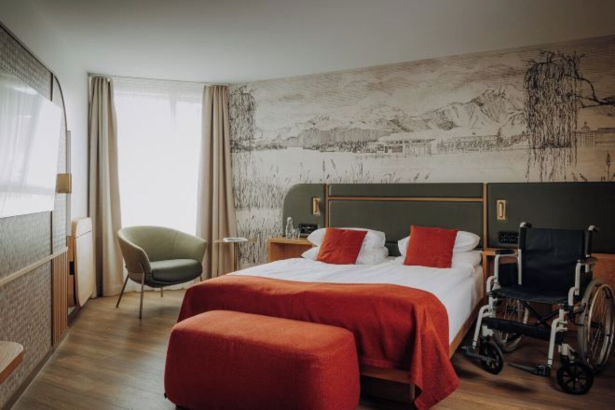 Quarto de hotel com cama, cadeira de rodas, poltrona verde e um quadro na parede.