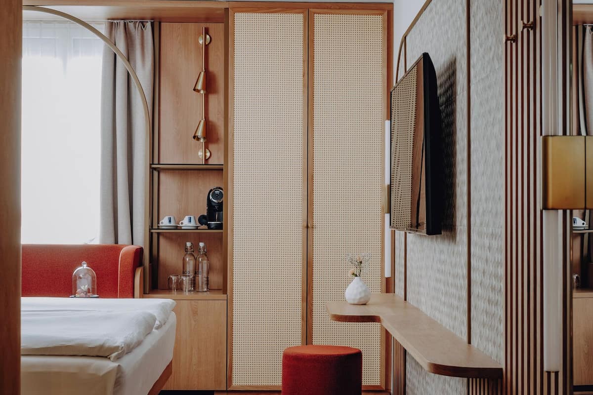 Quarto de hotel moderno com cama de madeira, um banquinho vermelho, escrivaninha de madeira, televisão e guarda-roupa com painel de encosto trançado.