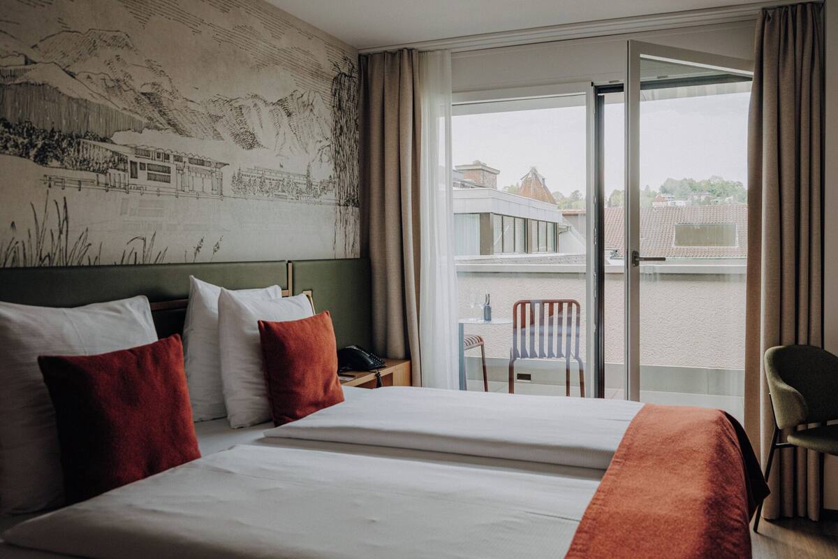 Quarto de hotel com cama, almofadas, escrivaninha com cadeira, janela com cortinas e vista para edifícios.