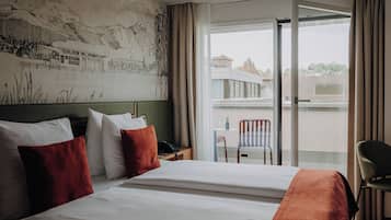 Quarto de hotel com cama, almofadas, escrivaninha com cadeira, janela com cortinas e vista para edifícios.