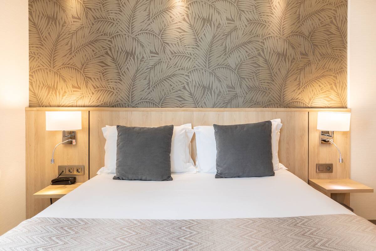 Quarto de hotel com uma cama grande, duas luminárias de cabeceira, um cabeceiro de madeira e um papel de parede estampado.