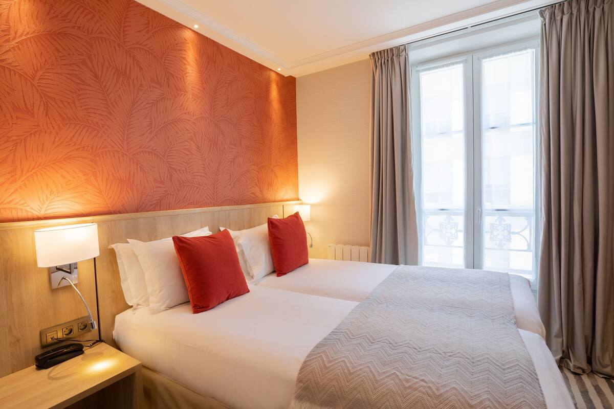 Quarto de hotel com cama grande, duas almofadas, mesa de cabeceira com abajur e janela com cortinas.