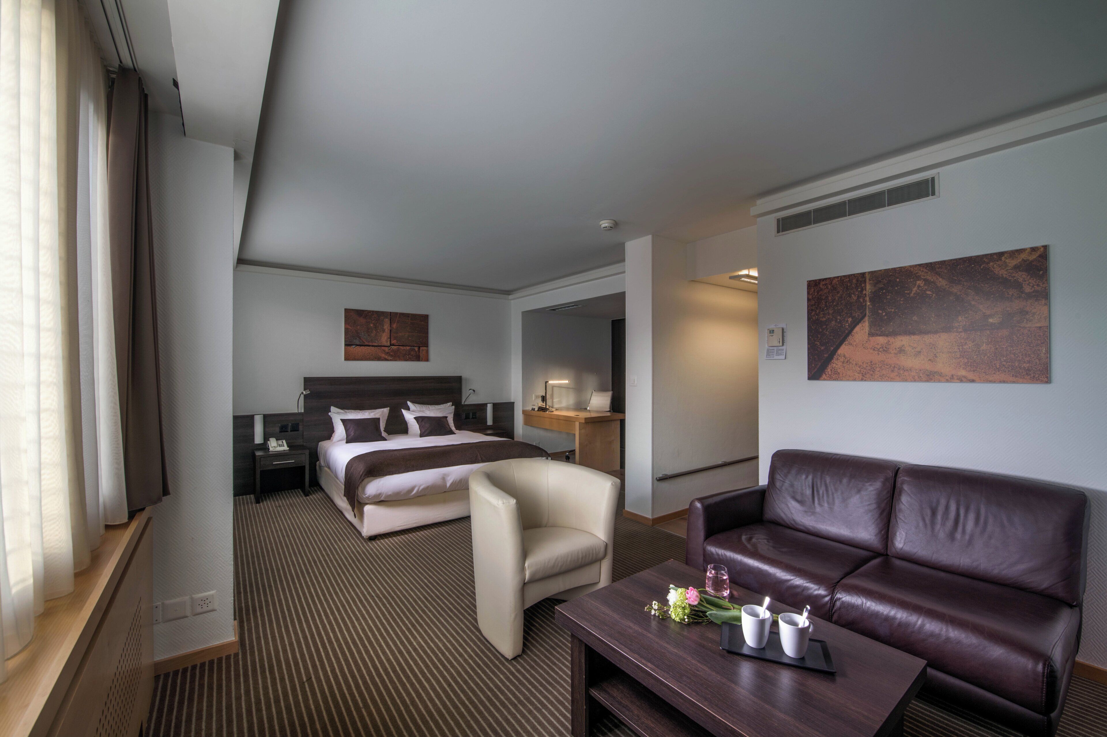 Quarto de hotel com cama, escrivaninha, sofá de couro e mesa de centro com bandeja de xícaras e flores.