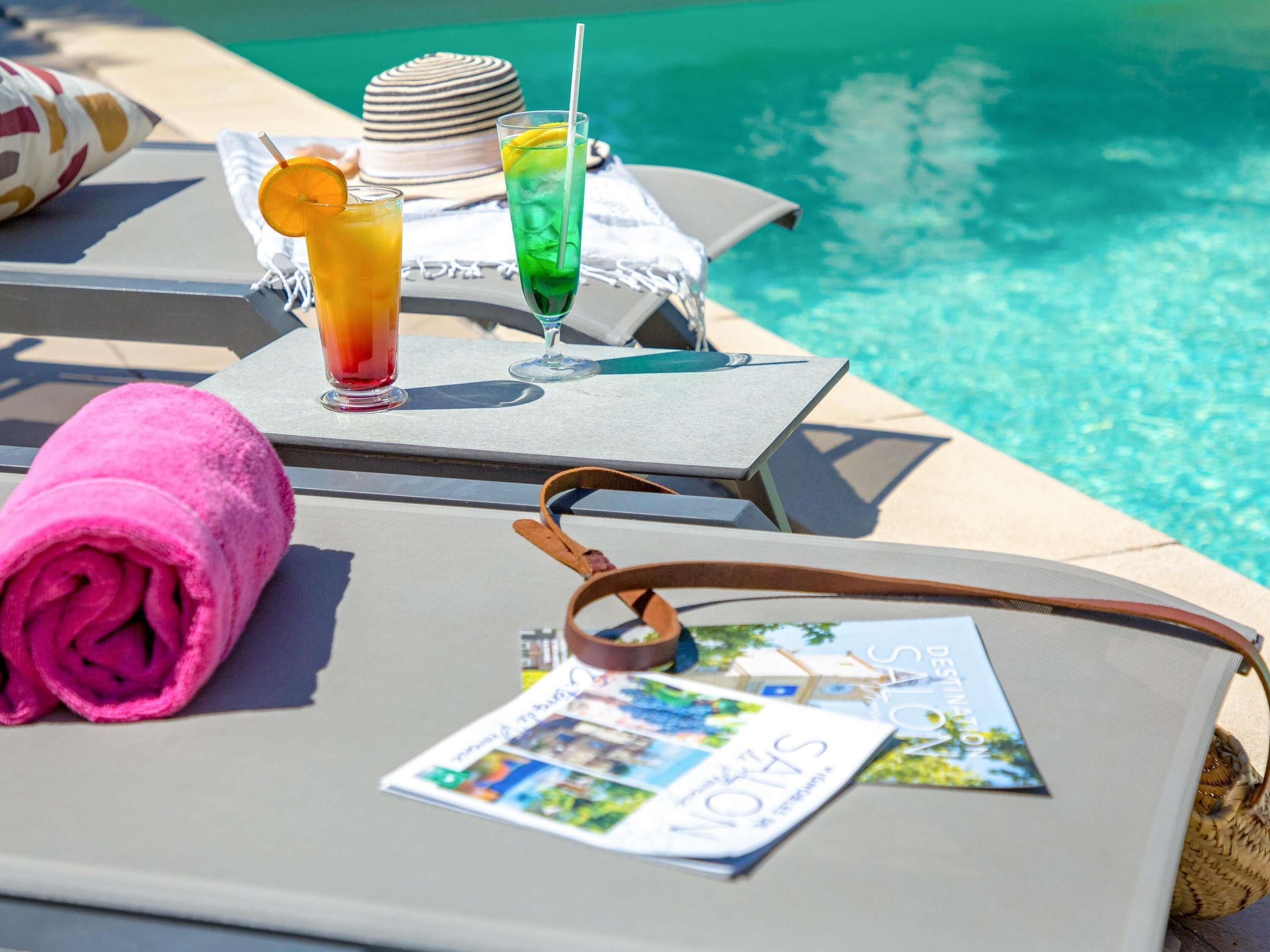 Uma área de descanso à beira da piscina, com espreguiçadeira, toalha enrolada, revista e duas bebidas sobre uma mesa.