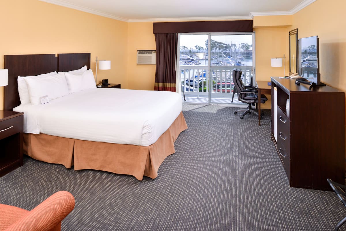 Quarto de hotel com uma cama grande, uma escrivaninha com cadeira, televisão e vista para uma marina através de uma porta de correr.