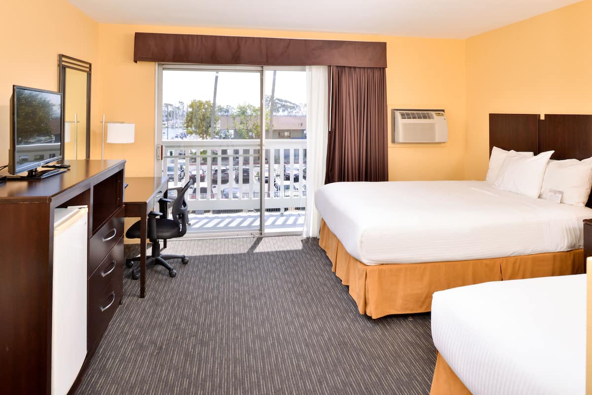 Quarto de hotel com uma cama, uma escrivaninha com cadeira, televisão e janela com vista para um parque de estacionamento e edifícios.