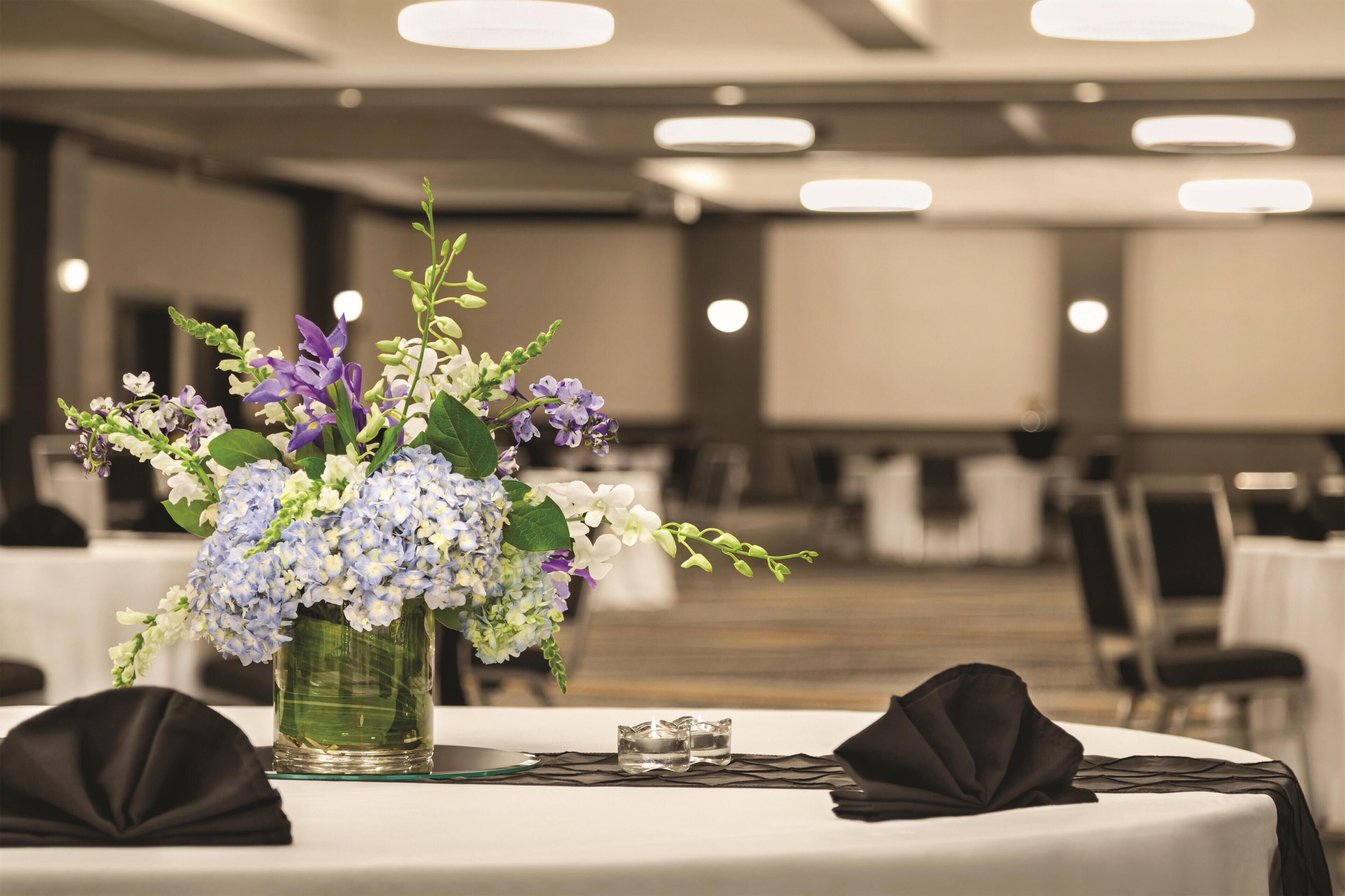 Uma mesa com arranjo floral, porta-guardanapos pretos e um copo de vidro transparente.