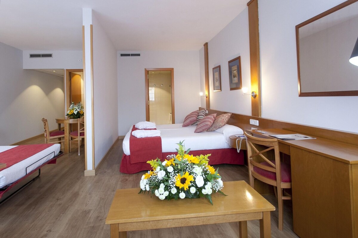 Quarto de hotel com cama, escrivaninha, cadeira, mesa com arranjo floral e espelho.