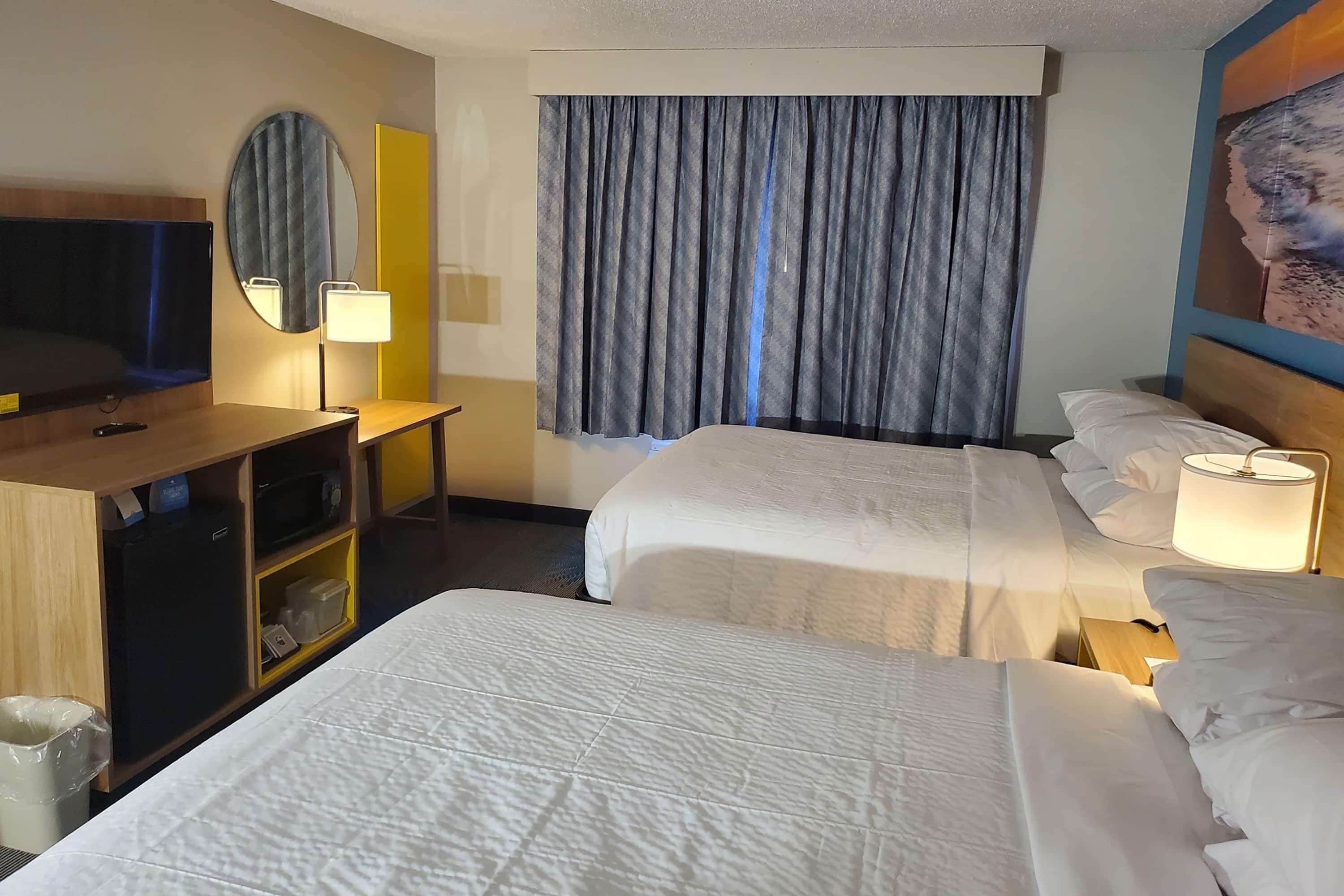 Quarto de hotel com duas camas, televisão, escrivaninha com abajur e janela com cortinas.