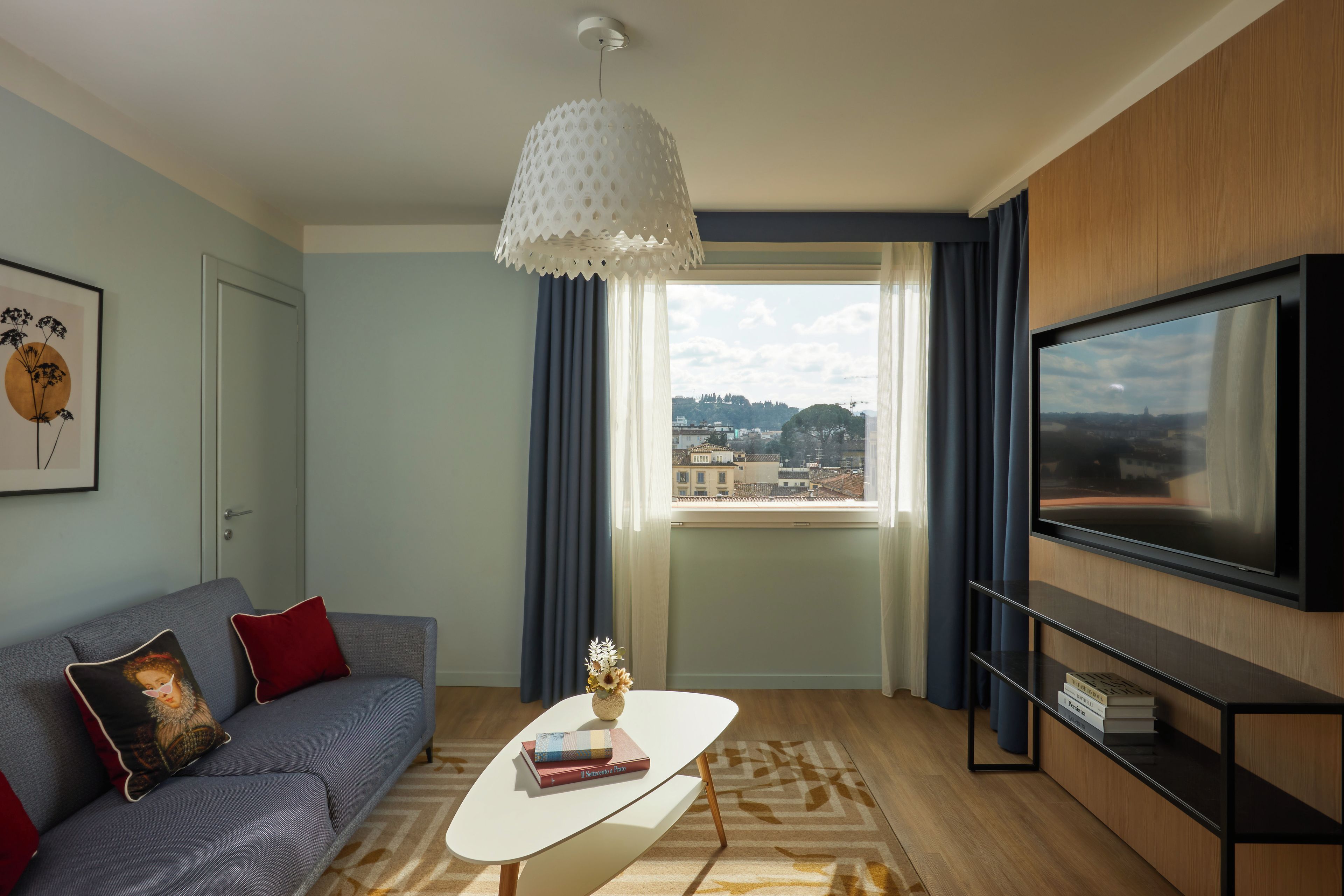 Uma sala de estar moderna com um sofá cinzento, uma mesa de centro branca, uma TV sobre um suporte de madeira e uma vista da cidade por uma grande janela.