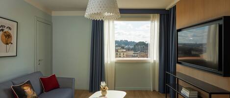 Uma sala de estar moderna com um sofá cinzento, uma mesa de centro branca, uma TV sobre um suporte de madeira e uma vista da cidade por uma grande janela.