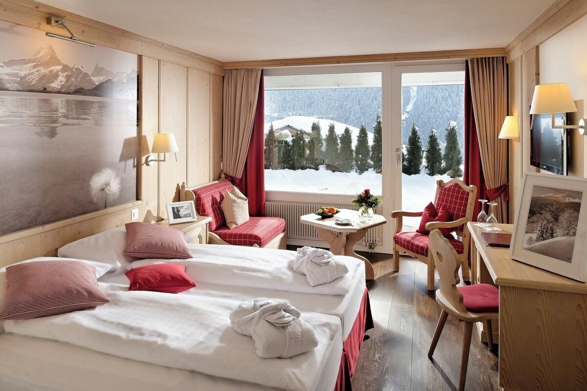 Quarto de hotel com duas camas, área de refeições e vista para uma montanha nevada.