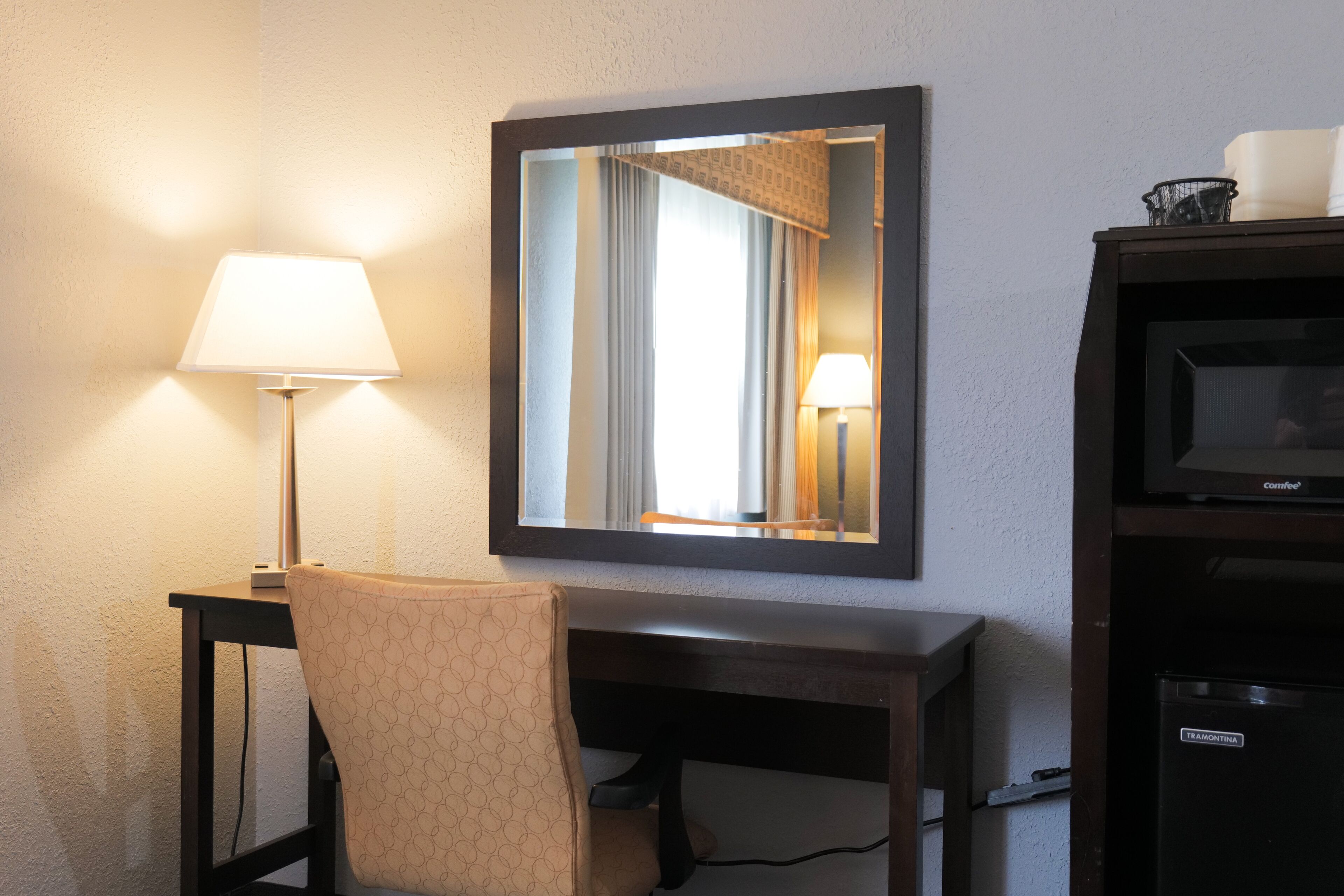Quarto de hotel com escrivaninha, cadeira, luminária, espelho e televisão.
