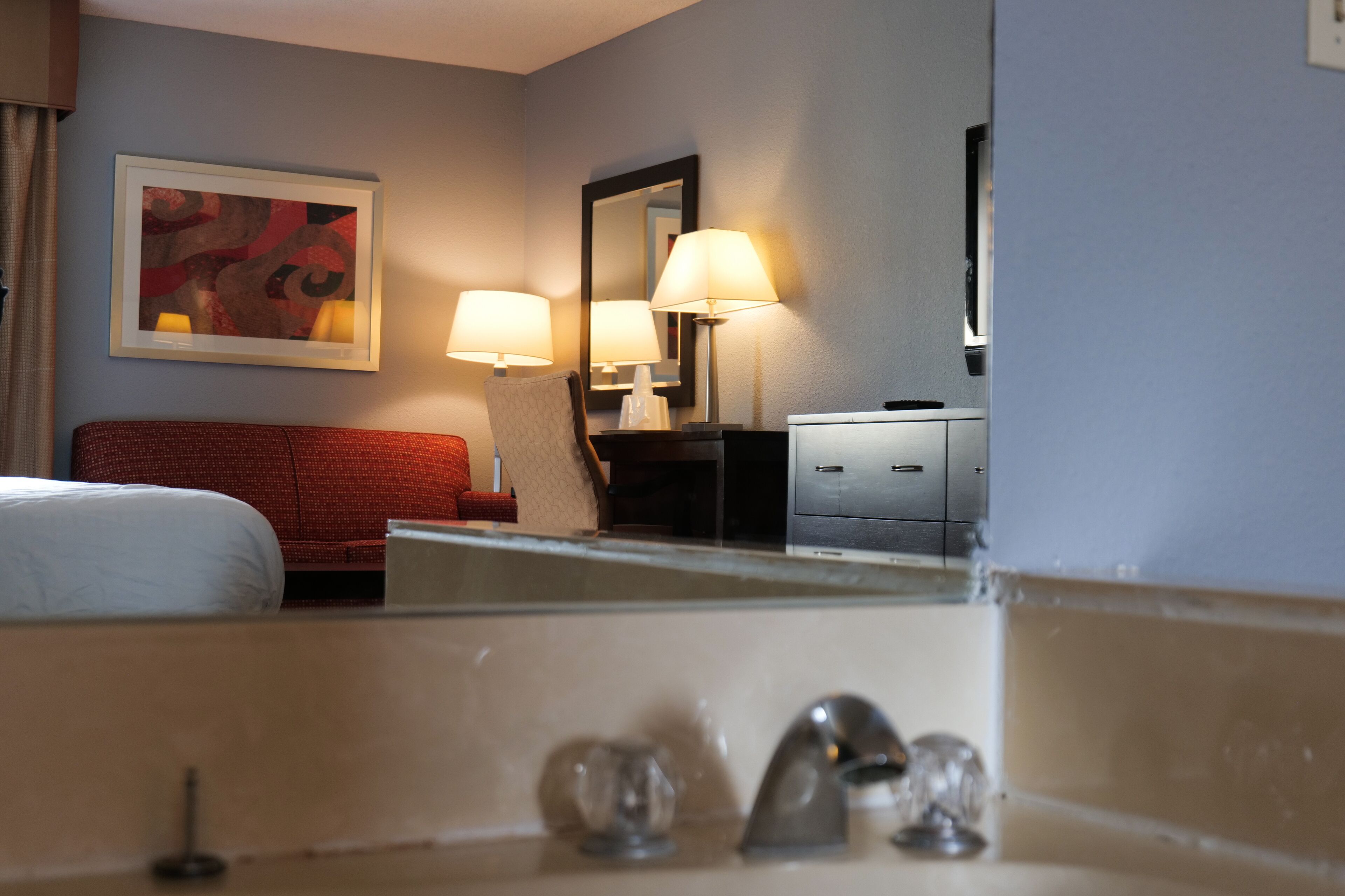 Quarto de hotel com uma cama, um sofá vermelho, uma escrivaninha com abajur, um espelho e um banheiro com pia e acessórios de vidro.