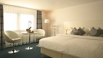Quarto de hotel com uma cama grande, duas cadeiras brancas, uma mesa redonda pequena e uma orquídea em vaso.