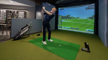 Um homem pratica swings de golfe numa sala com um tapete verde, uma bolsa de golfe e um ecrã a exibir um campo de golfe.