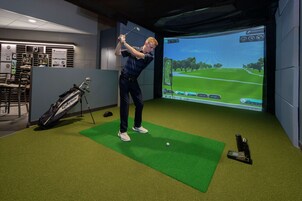 Um homem pratica swings de golfe numa sala com um tapete verde, uma bolsa de golfe e um ecrã a exibir um campo de golfe.