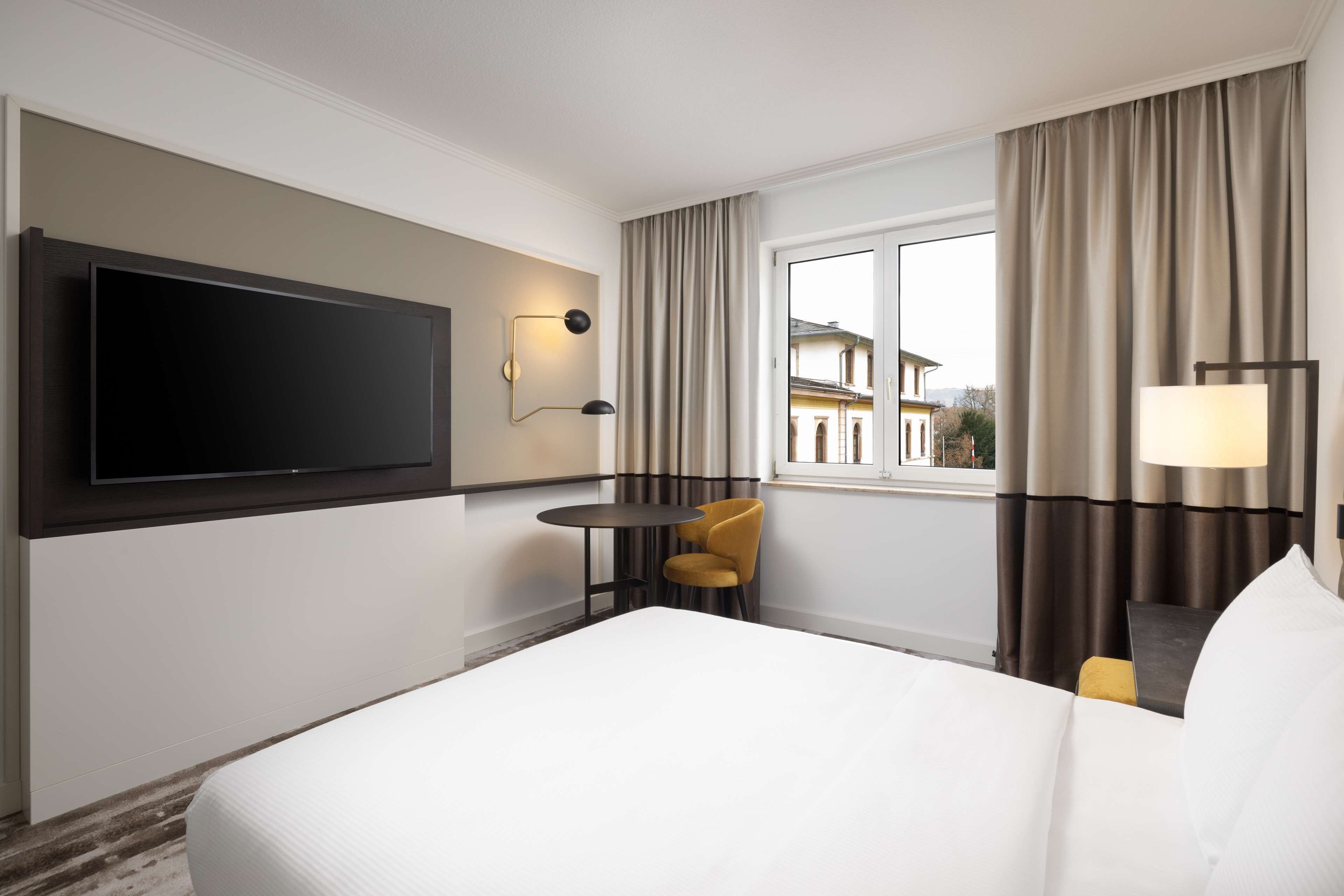 Quarto de hotel com uma cama grande, uma televisão de tela plana na parede, uma mesa redonda pequena com uma cadeira e uma janela com cortinas.