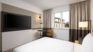 Quarto de hotel com uma cama grande, uma televisão de tela plana na parede, uma mesa redonda pequena com uma cadeira e uma janela com cortinas.