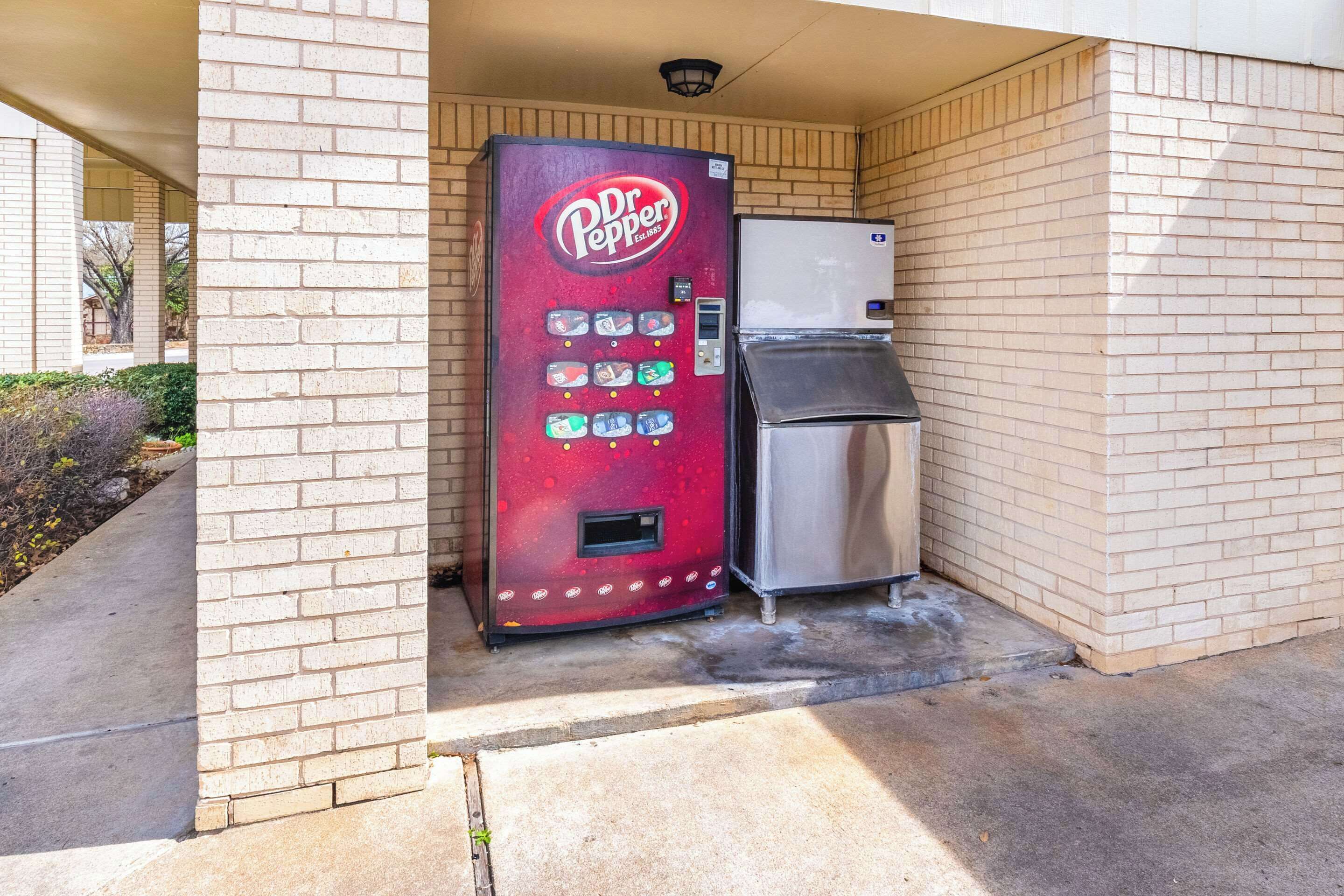 Máquina de refrigerantes Dr. Pepper ao lado de uma geladeira em área externa coberta, com paredes de tijolo.