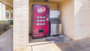 Máquina de refrigerantes Dr. Pepper ao lado de uma geladeira em área externa coberta, com paredes de tijolo.